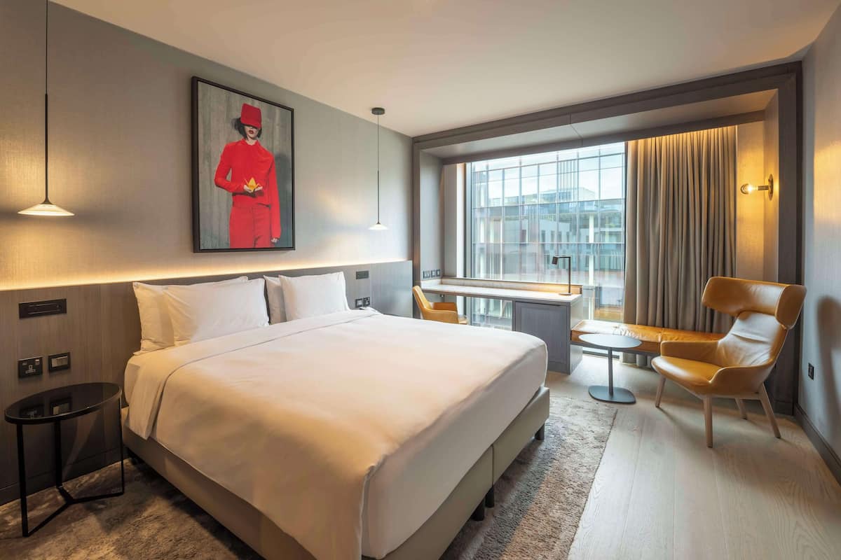 Quarto de hotel com uma cama grande, uma escrivaninha, uma cadeira, uma mesa pequena e uma janela ampla com cortinas.
