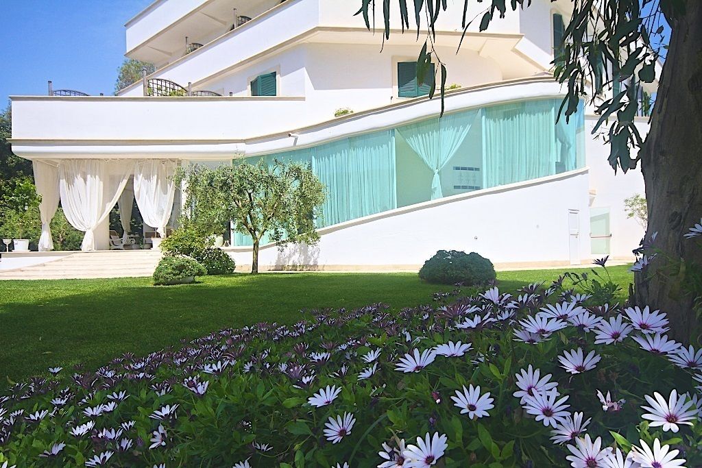 Um edifício moderno de dois andares com fachada branca, uma grande janela de vidro e um jardim bem cuidado com flores roxas.