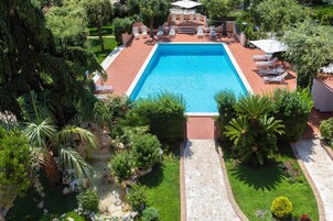 Uma piscina retangular com um deck ladrilhado, rodeada por espreguiçadeiras e guarda-sóis, com um jardim e árvores ao fundo.
