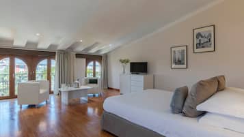 Quarto espaçoso com uma cama grande, uma escrivaninha e uma cadeira. Há duas janelas com cortinas, uma TV em um móvel e uma obra de arte na parede.