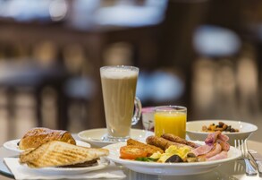 Disposição para o café da manhã com diversos pratos, incluindo torradas, ovos, linguiça e um copo de suco de laranja.