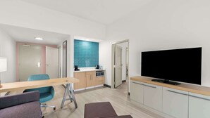 Quarto de hotel moderno com escrivaninha de madeira, cadeira azul, televisão de tela plana e cozinha compacta.