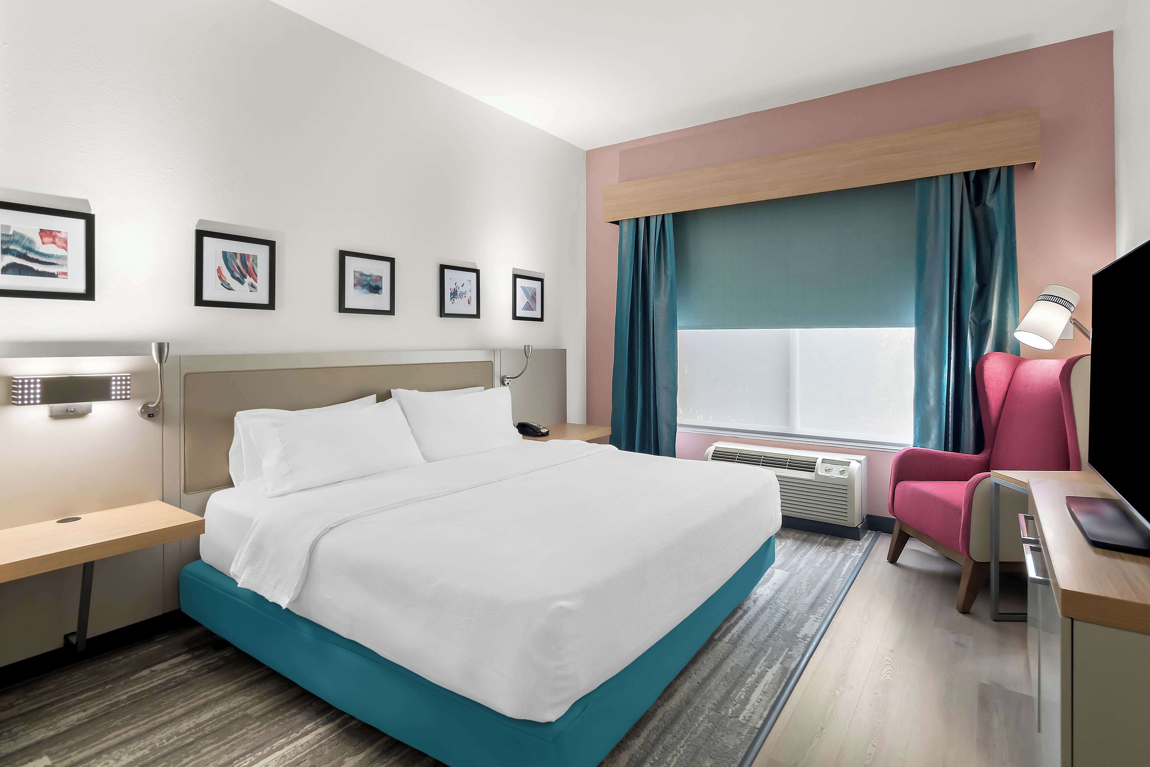Quarto de hotel com cama, escrivaninha, cadeira, televisão e quadros na parede.