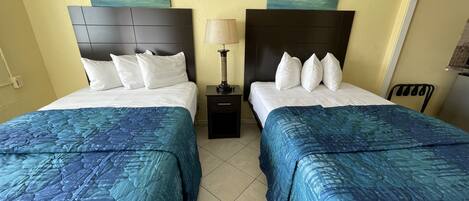 Quarto de hotel com duas camas, cada uma com cabeceira e almofadas brancas. Há duas pinturas na parede retratando barcos no mar.