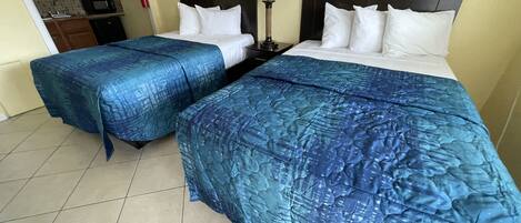 Quarto de hotel com duas camas, cada uma com colcha azul acolchoada, luminária de cabeceira e cabeceira branca.