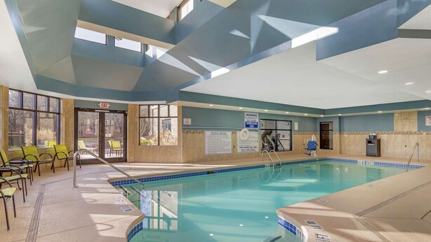Piscina interior com água cristalina, escada de piscina e cadeira de salva-vidas. Há cadeiras de descanso e um aviso sobre os riscos da piscina.