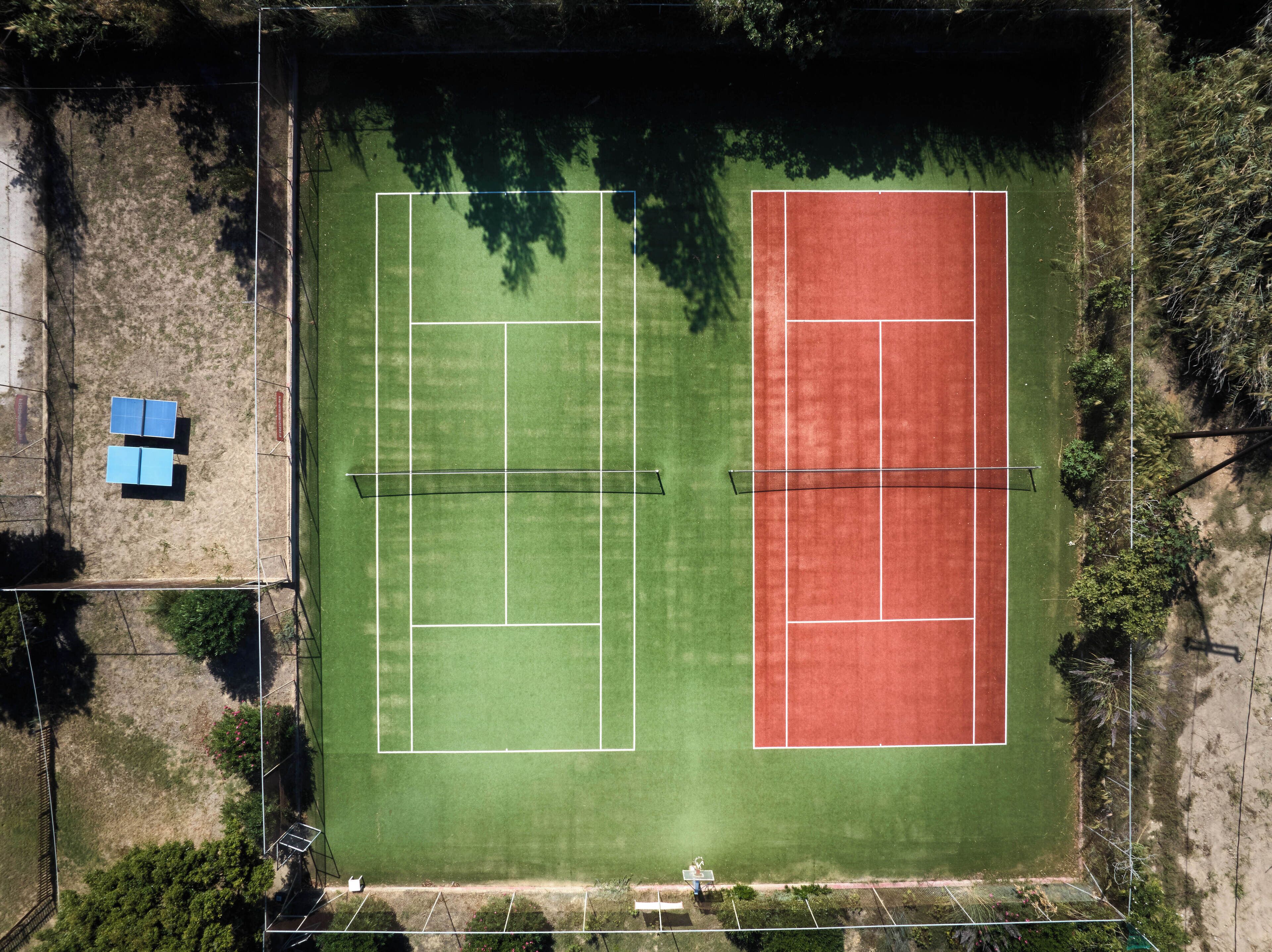 Vista aérea de uma quadra de tênis com duas quadras vizinhas, cercada por árvores e um pequeno edifício com painéis solares.