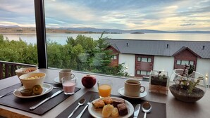 Mesa de café da manhã com vista panorâmica para um lago, incluindo uma casa e árvores.