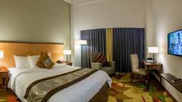Quarto de hotel com uma cama grande, duas mesas de cabeceira com abajures, uma cadeira, uma escrivaninha e uma televisão fixada na parede.