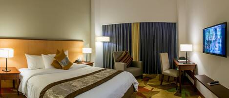 Quarto de hotel com uma cama grande, duas mesas de cabeceira com abajures, uma cadeira, uma escrivaninha e uma televisão fixada na parede.