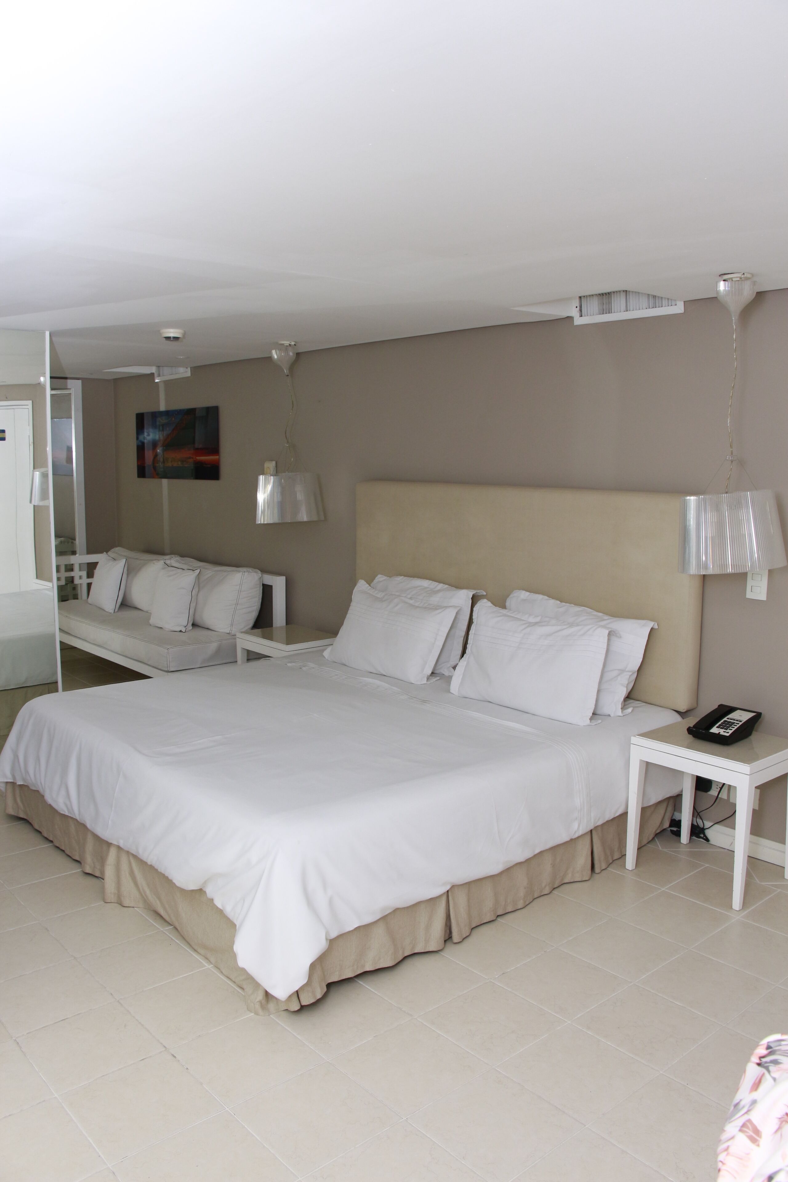 Quarto de hotel com uma cama grande, mesa de cabeceira com telefone, pequena mesa com abajur e um quadro na parede.