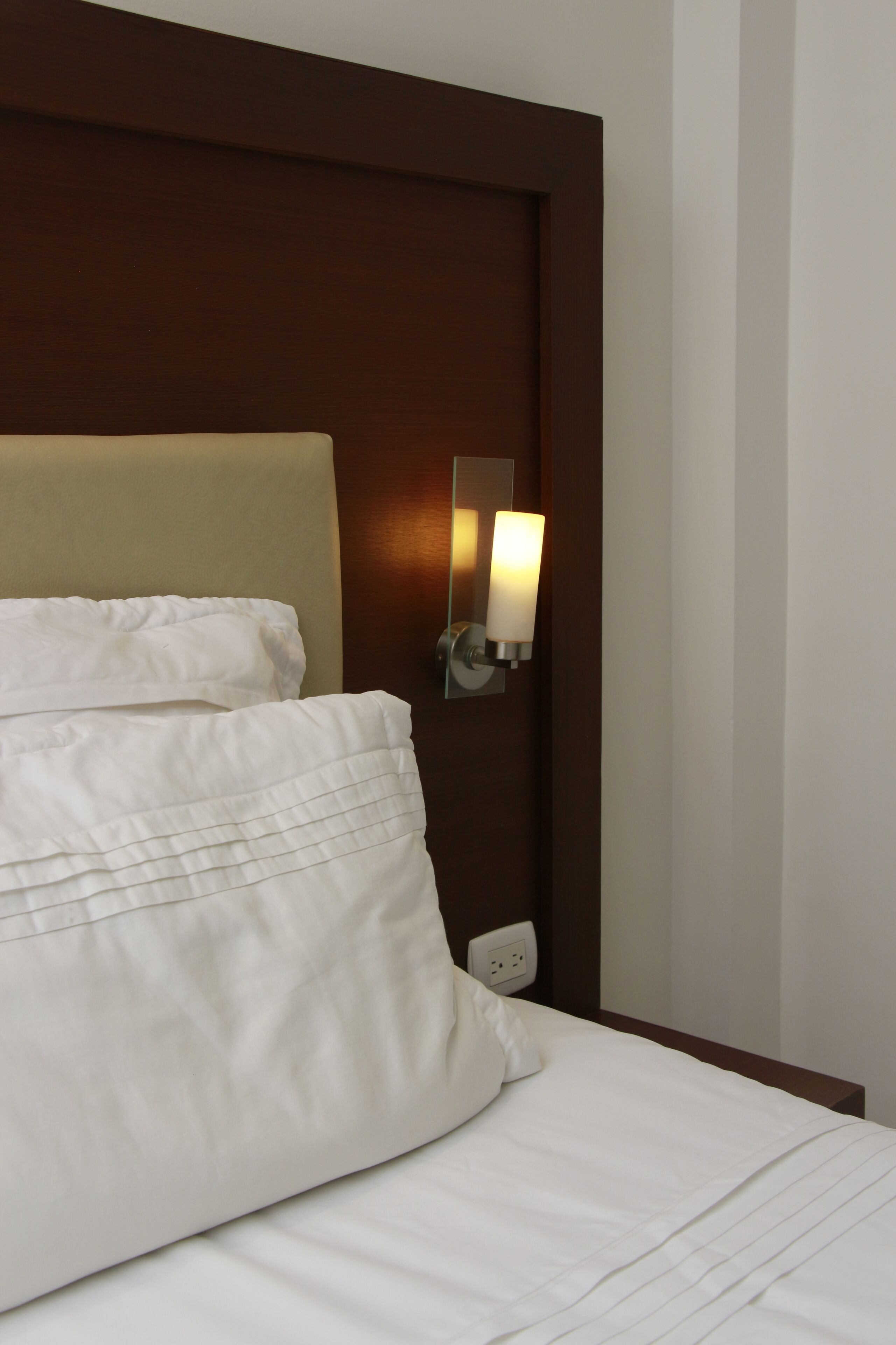 Quarto de hotel com uma cama bem arrumada, luminária na parede e uma tomada.