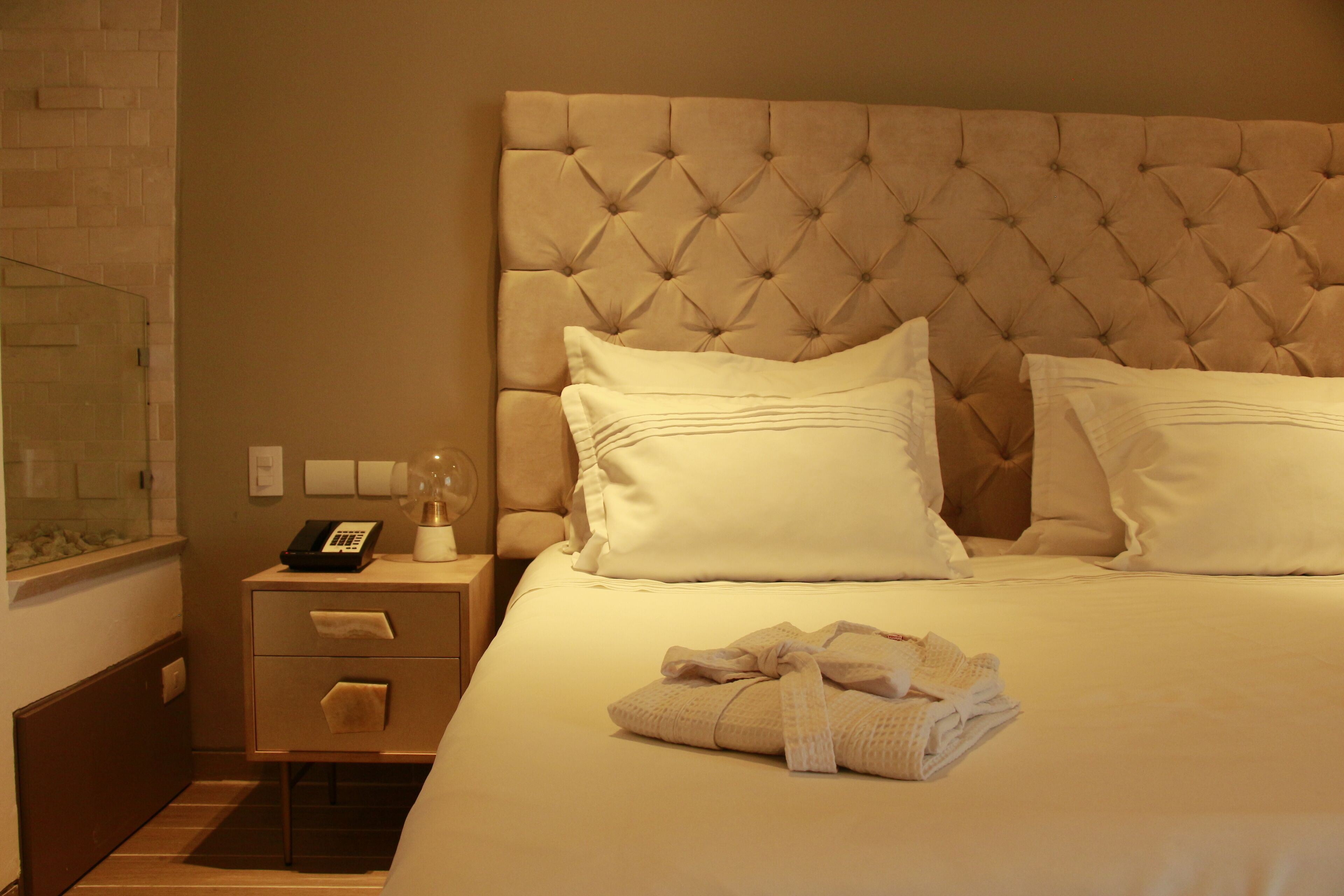 Quarto de hotel com cabeceira estofada, roupa de cama branca, mesa de cabeceira com telefone e toalha dobrada na cama.