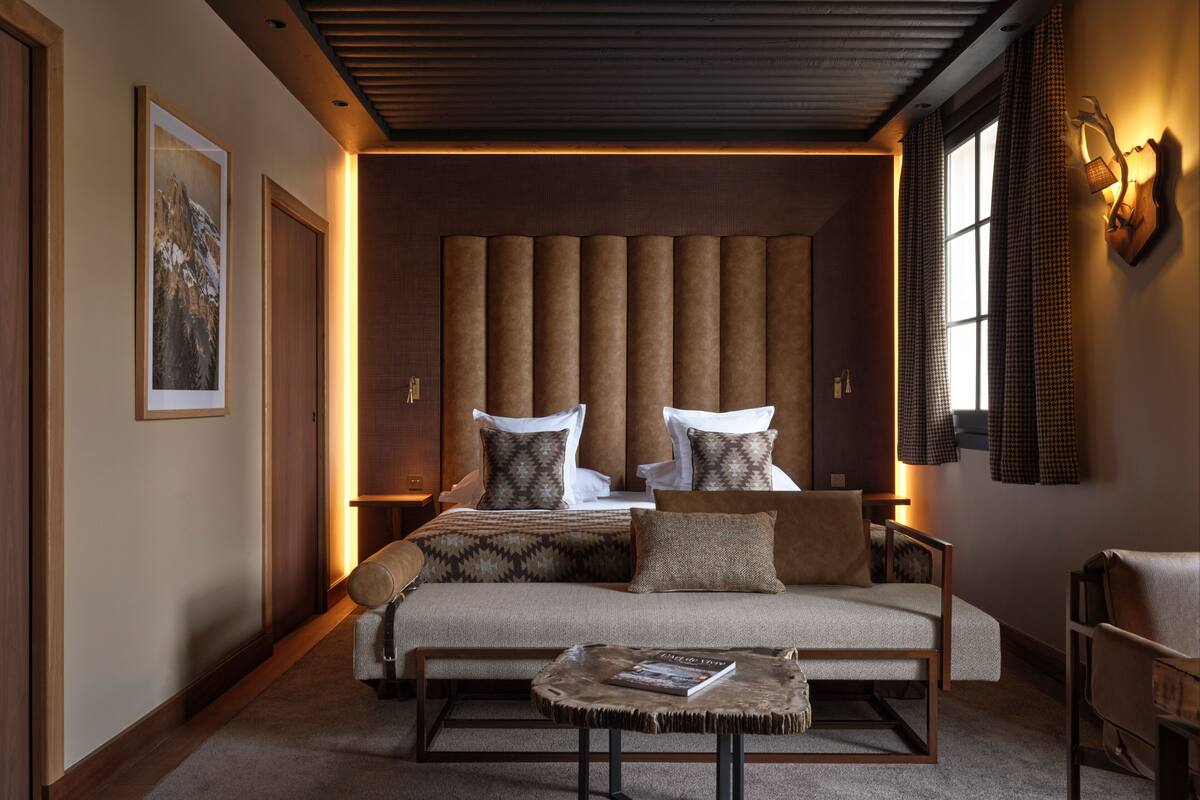 Quarto de hotel moderno com uma cama grande, um sofá, uma mesa pequena e uma luminária fixada na parede.