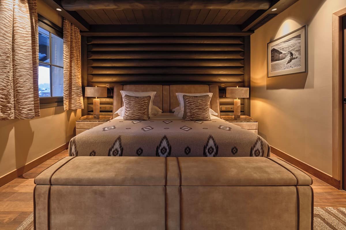 Quarto com teto de madeira, uma cama grande com roupa de cama estampada, duas luminárias de cabeceira e um quadro na parede.