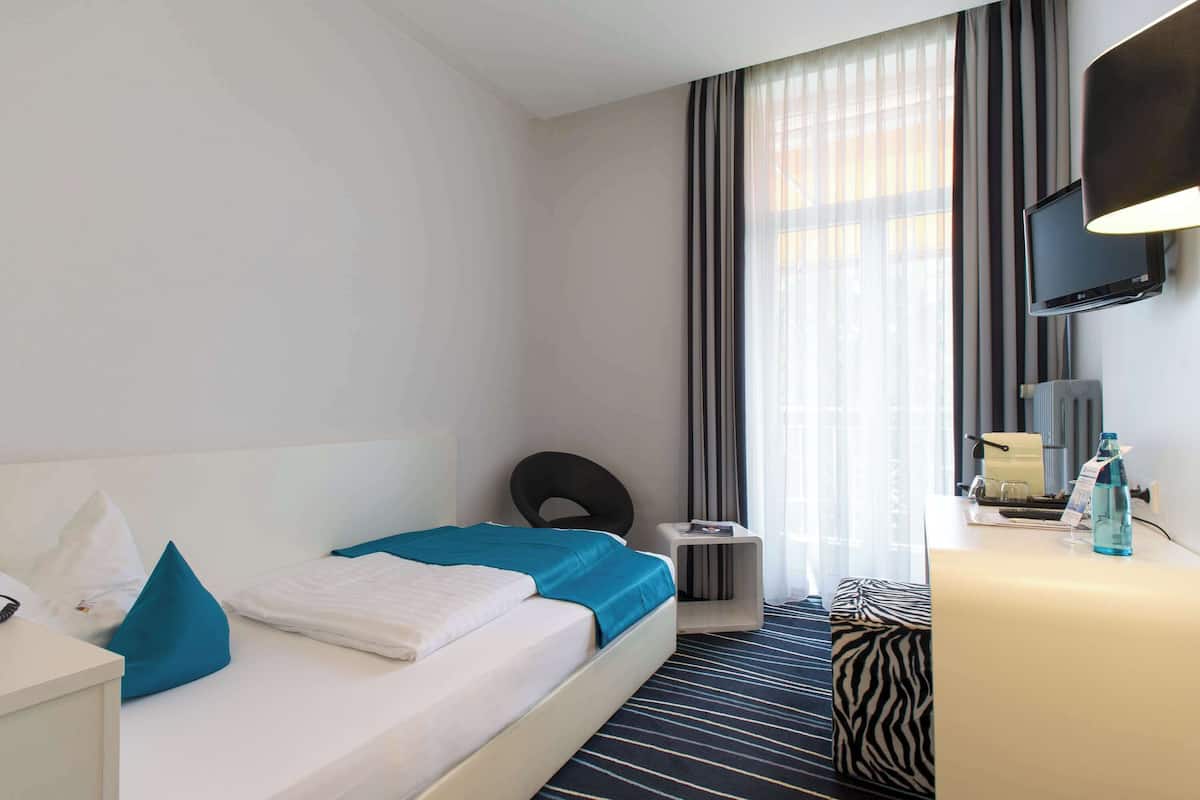 Quarto de hotel com cama, escrivaninha, cadeira, televisão e janela com cortinas.