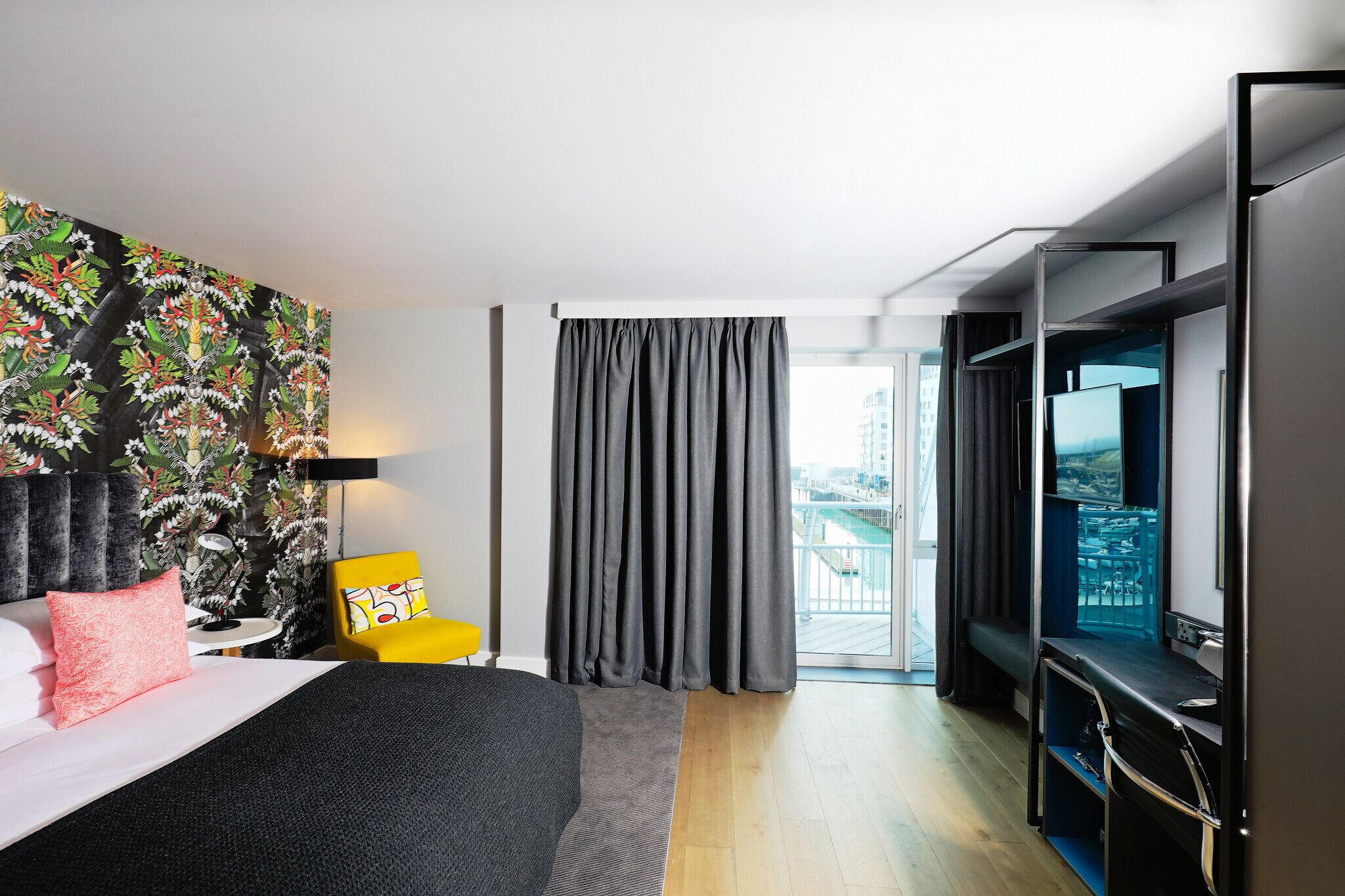 Quarto de hotel moderno com uma cama grande, uma escrivaninha com televisão de tela plana, varanda com vista para a cidade e uma parede floral vibrante.