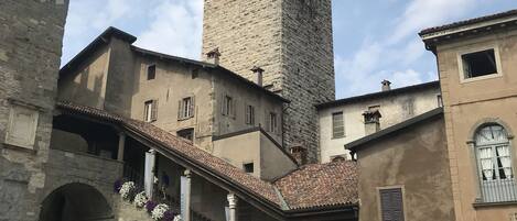 Uma praça histórica com uma imponente torre do relógio, edifícios de pedra e área pavimentada para pedestres.