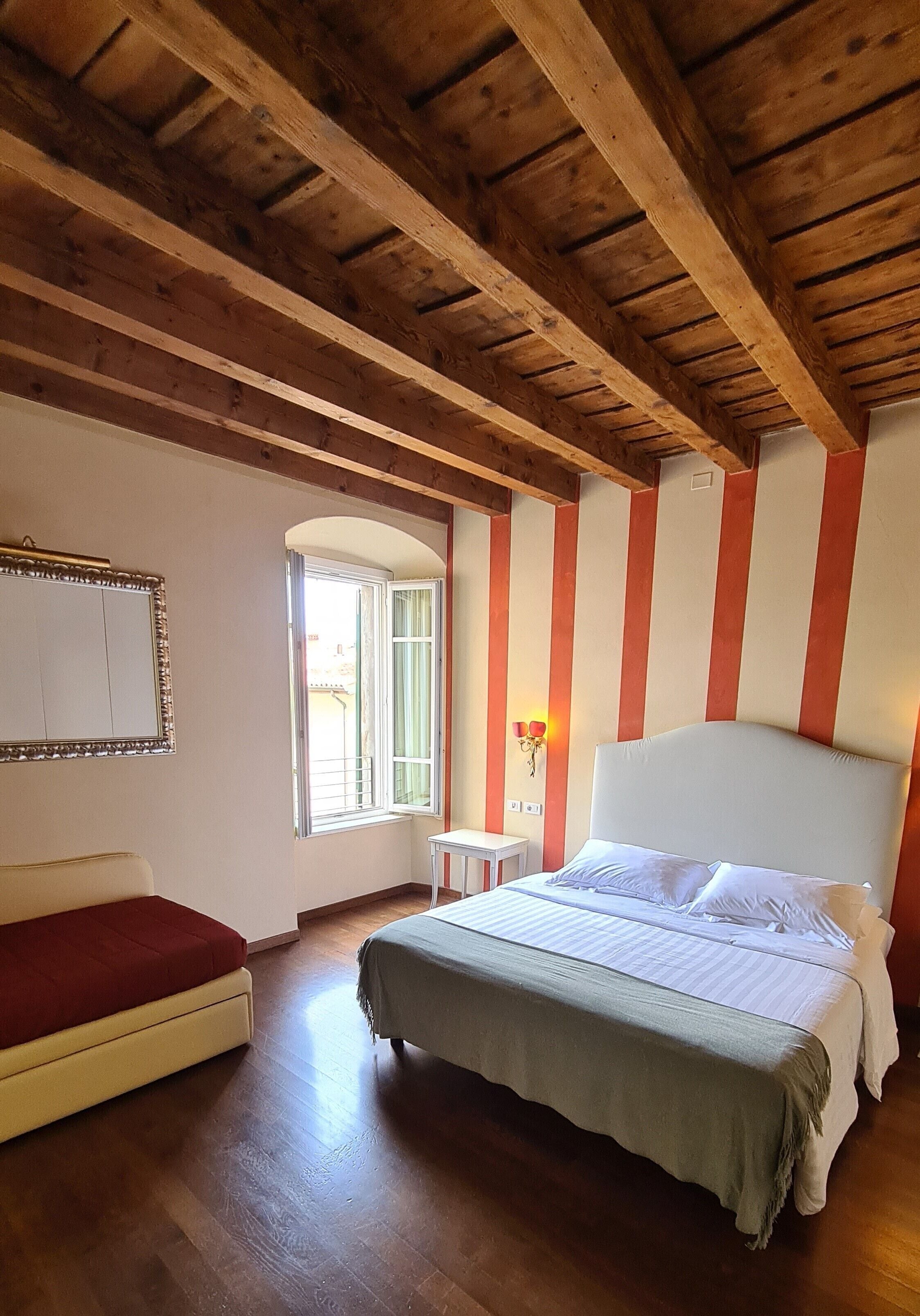 Quarto com teto de madeira, cama com roupa de cama branca, sofá vermelho, mesa pequena e janela com cortinas.