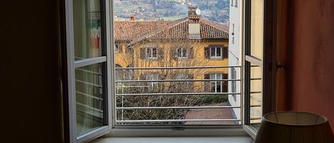 Quarto com janela para uma paisagem montanhosa, uma luminária em uma mesa de cabeceira e vista de um edifício com telhado de azulejos.