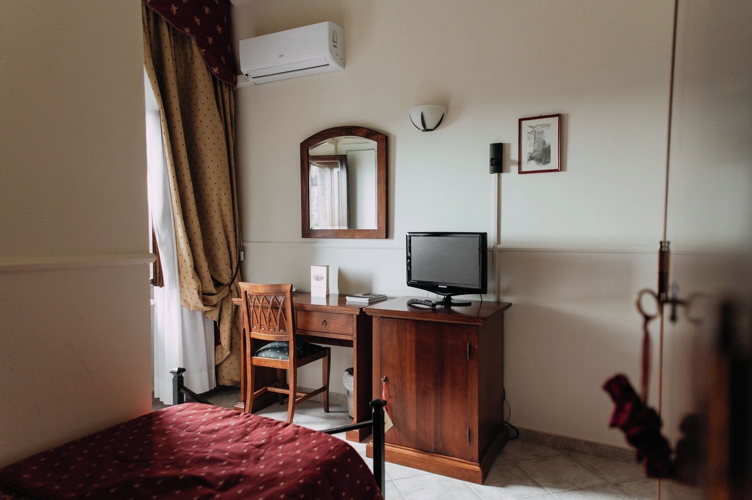 Quarto de hotel com cama, escrivaninha, cadeira, televisão, guarda-roupa, espelho e ar condicionado.