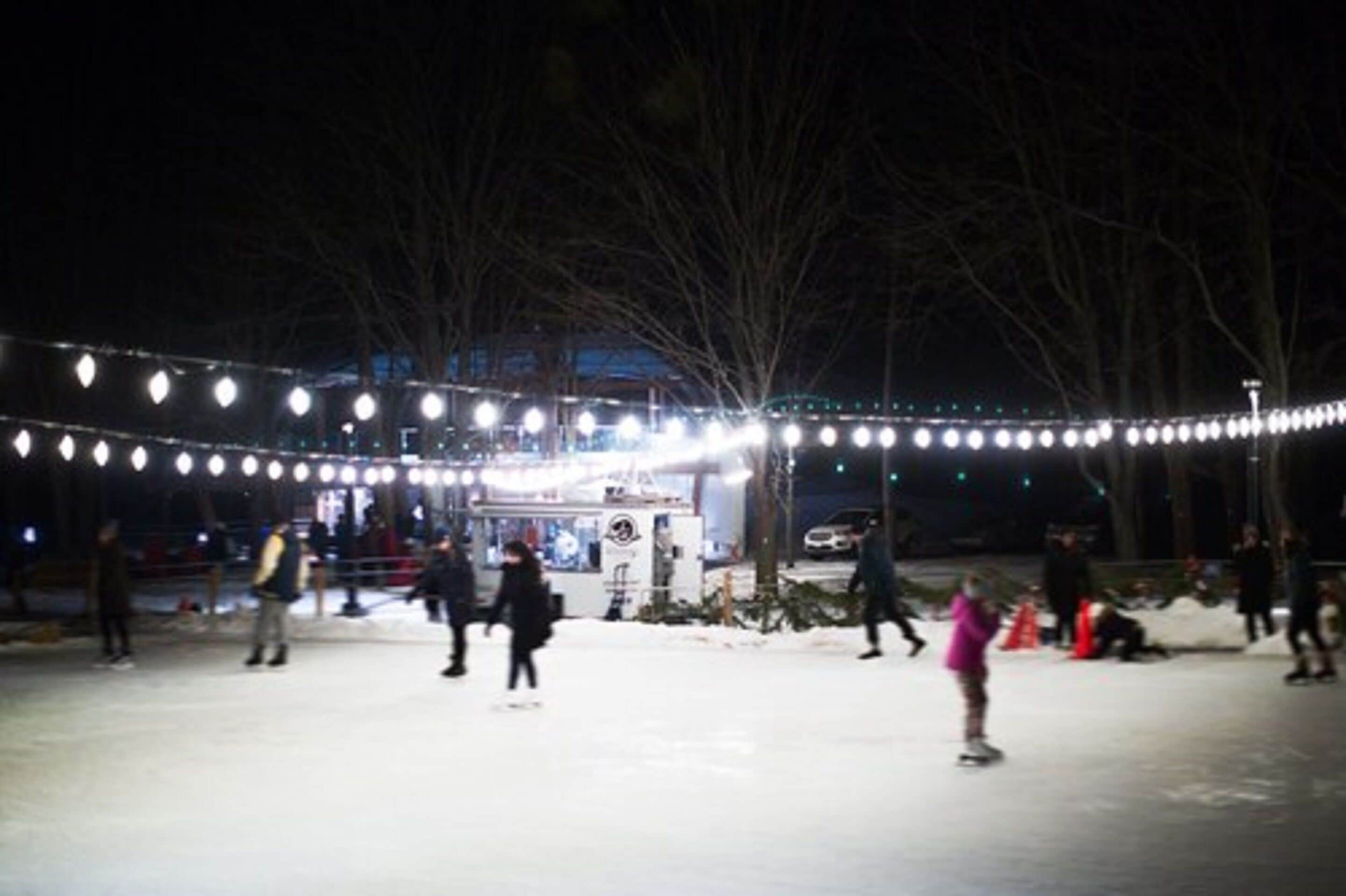 Uma pista de patinação no gelo ao ar livre à noite, com pessoas patinando e um quiosque de comida ao fundo.