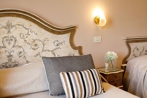 Quarto com cama, duas almofadas, mesa de cabeceira com um vaso de flores e luminária fixada na parede.