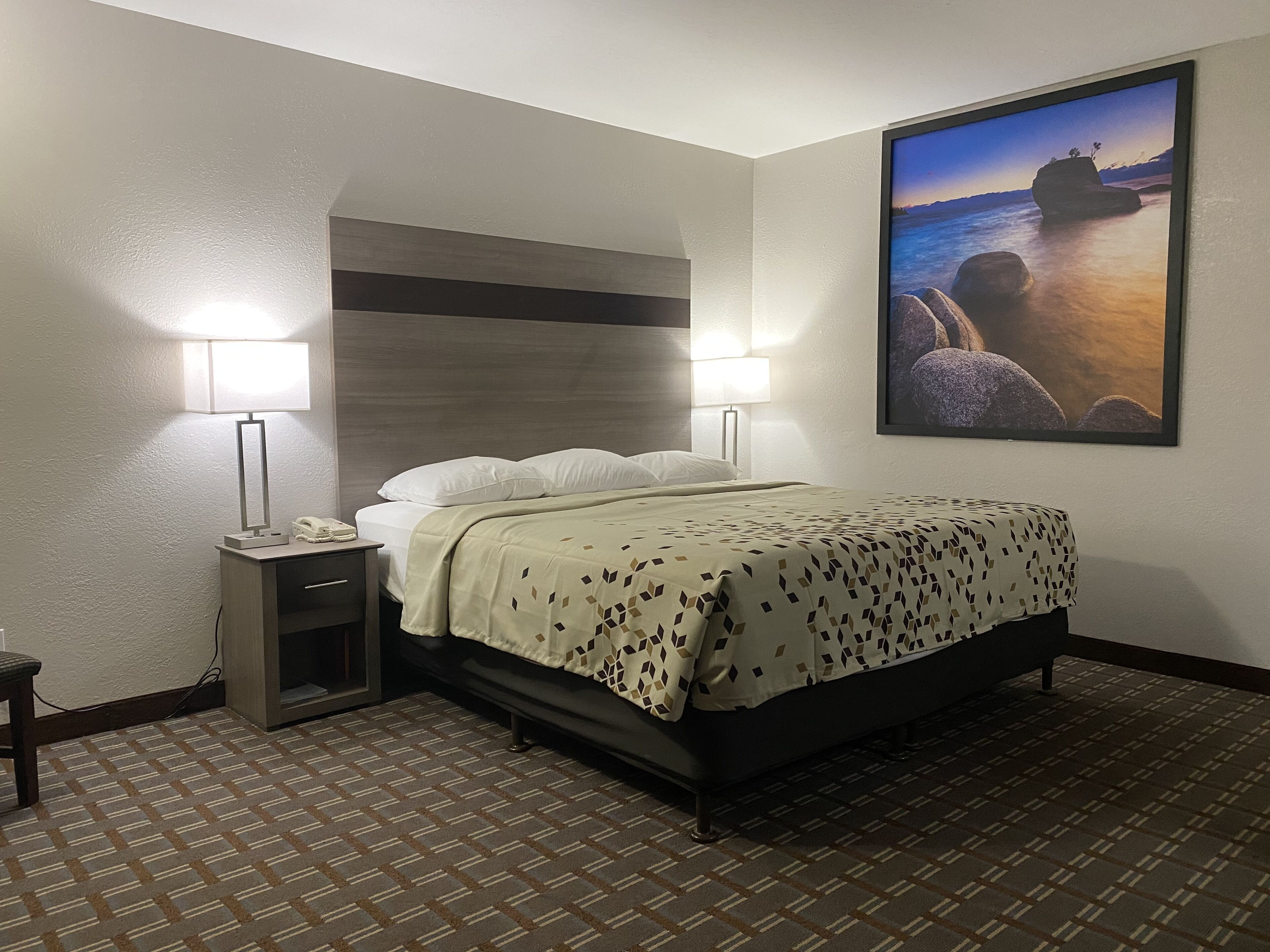 Quarto de hotel com uma cama, duas luminárias de cabeceira, mesa de cabeceira, telefone e um quadro com uma cena de praia.