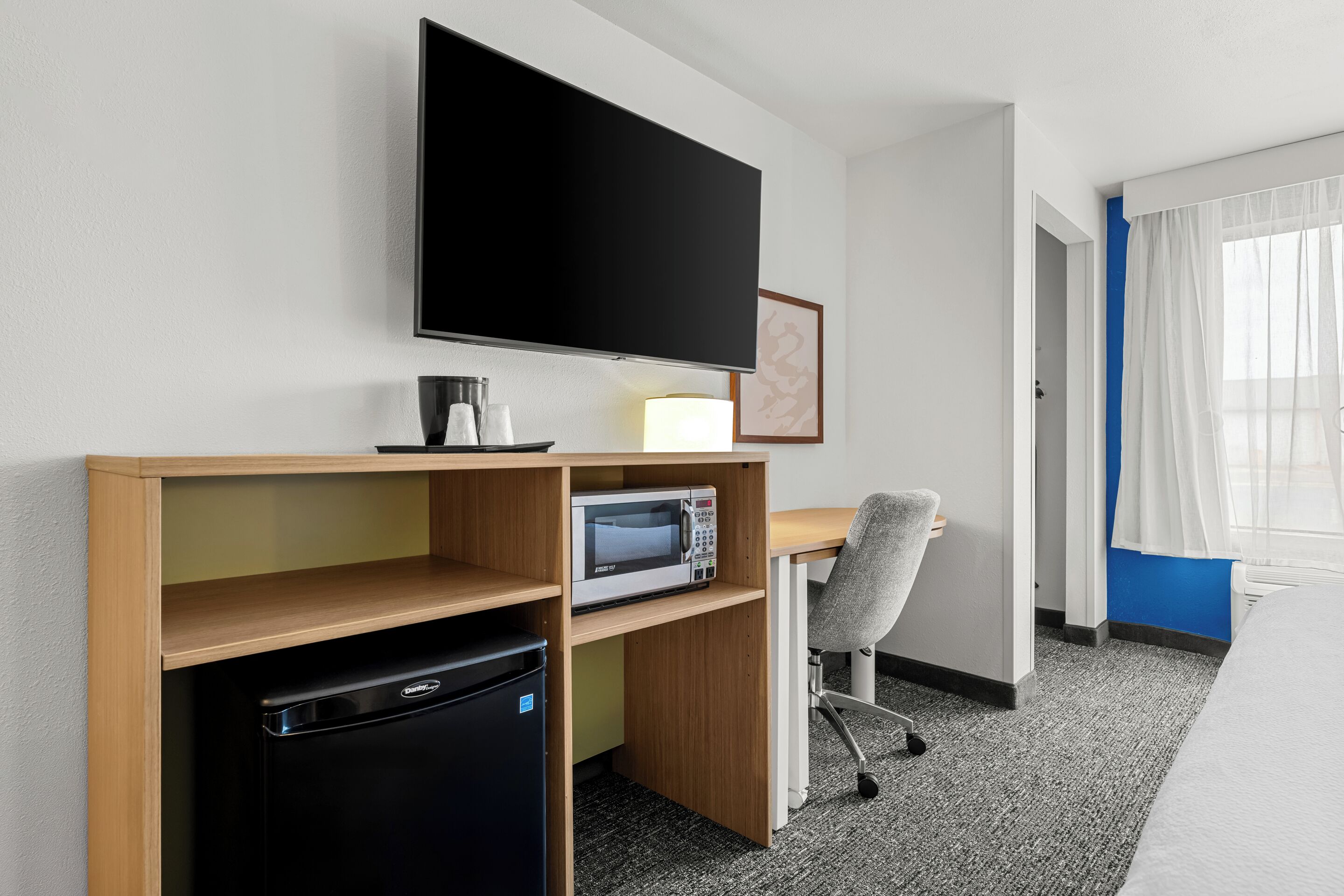 Quarto de hotel com televisão de tela plana, micro-ondas, mini-geladeira, escrivaninha, cadeira e cama.