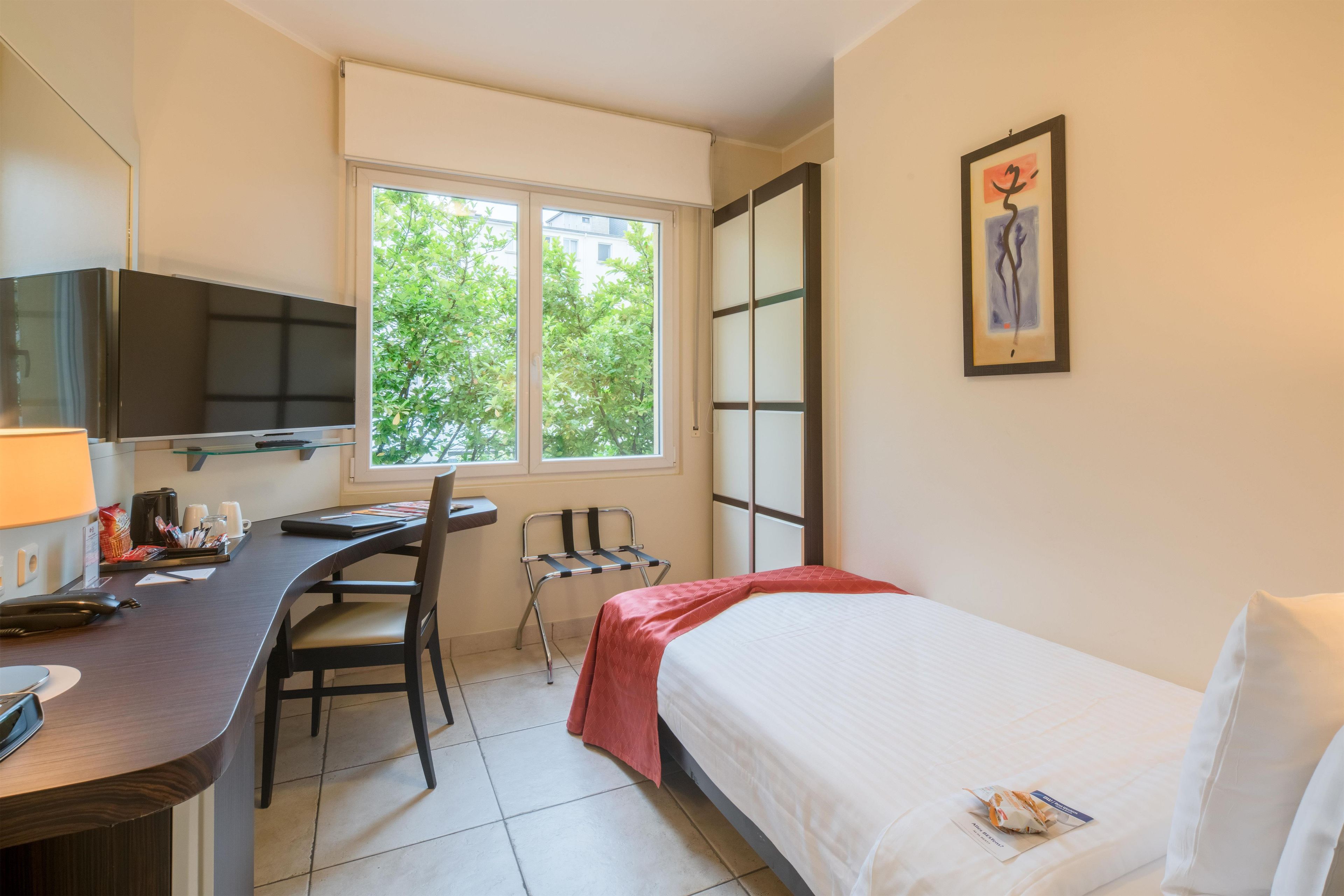 Quarto de hotel com cama, escrivaninha com cadeira, televisão e janela com vista para árvores.