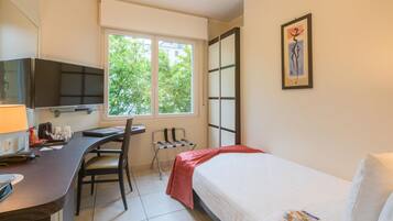 Quarto de hotel com cama, escrivaninha com cadeira, televisão e janela com vista para árvores.