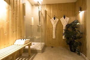 Quarto com painéis de madeira, chuveiro em cabine de vidro, um banco com toalhas e dois roupões brancos pendurados na parede.
