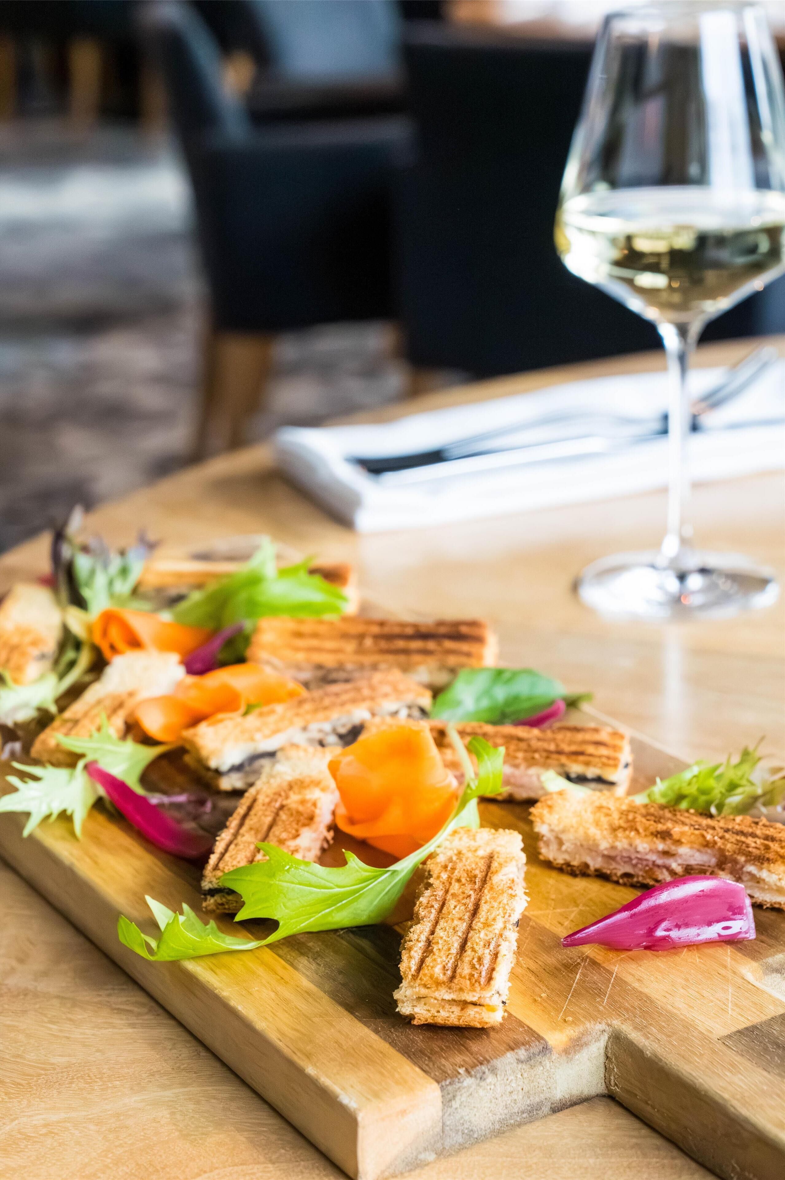 Uma tábua de madeira com sanduíches e um copo de vinho branco sobre uma mesa.