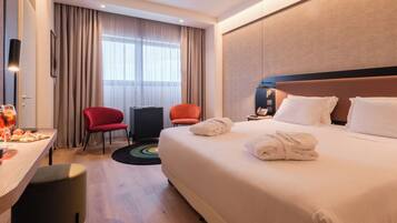 Quarto de hotel com uma cama grande, duas cadeiras, uma escrivaninha com abajur e vista para o exterior pela janela.