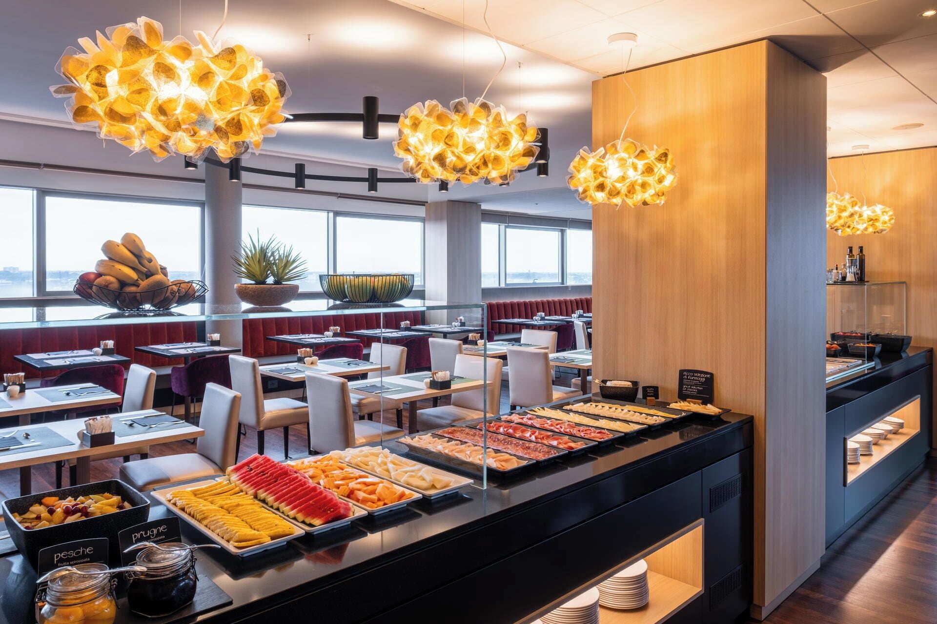 Uma área de buffet com diversas opções de comida, um espaço de estar com mesas e cadeiras, e uma janela grande com vista.