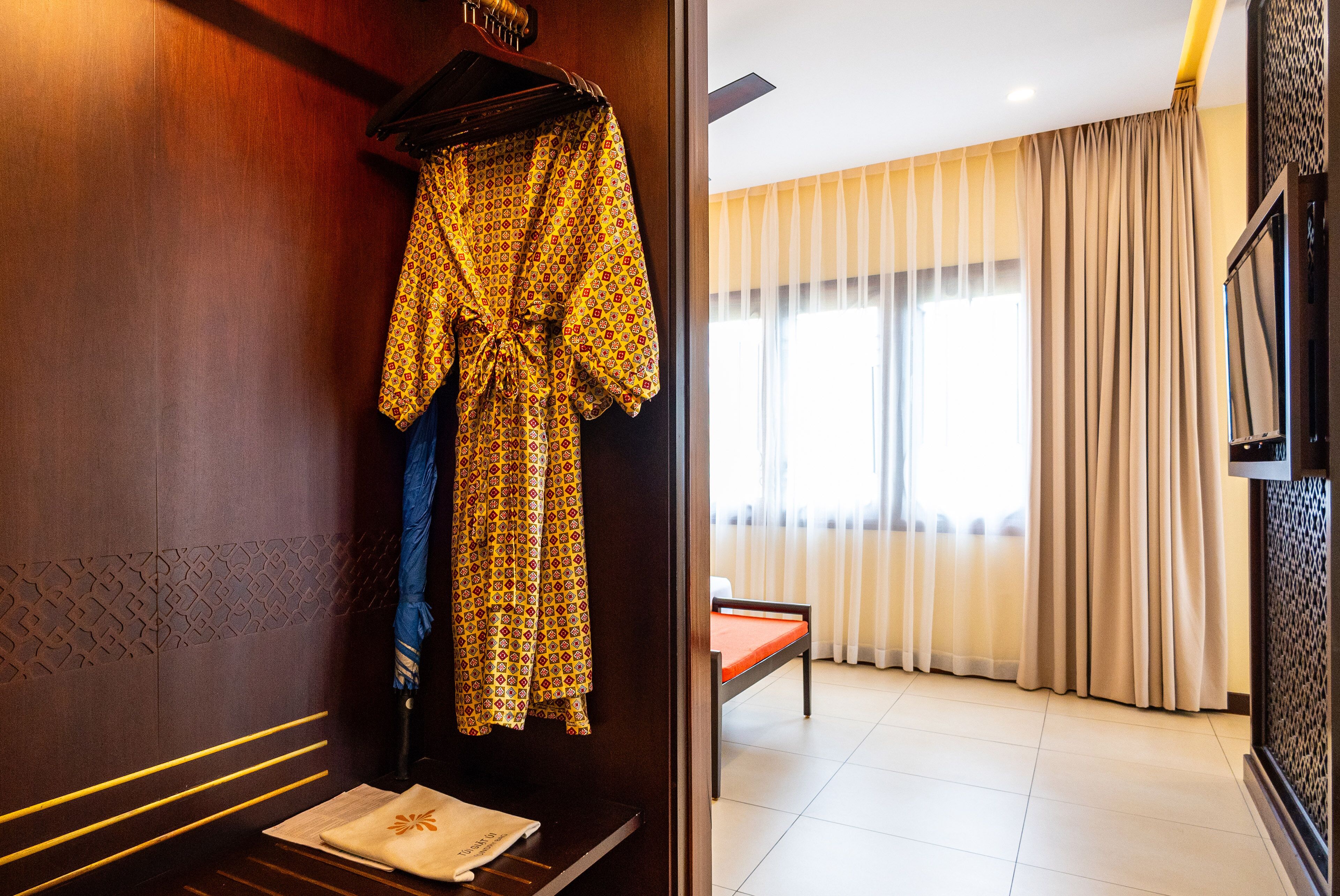Quarto de hotel com guarda-roupa de madeira, cama com colchão laranja, janela com cortinas bege e televisão na parede.