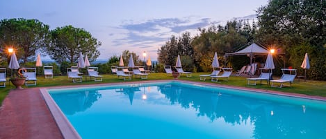 Piscina com espreguiçadeiras e guarda-sóis, cercada por árvores e um céu claro.