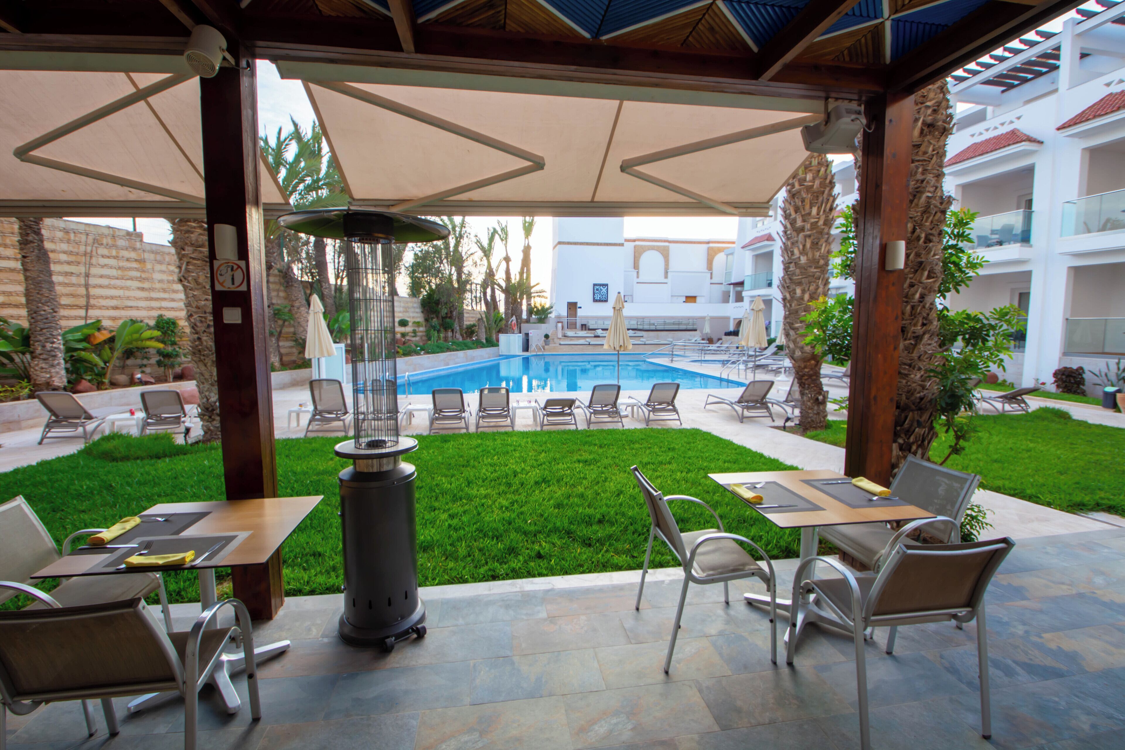 Uma área de piscina ao ar livre com espreguiçadeiras e mesas, cercada por vegetação e um edifício ao fundo.
