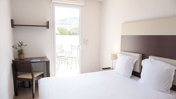 Quarto de hotel com cama, escrivaninha, cadeira, luminária e vista para o exterior.
