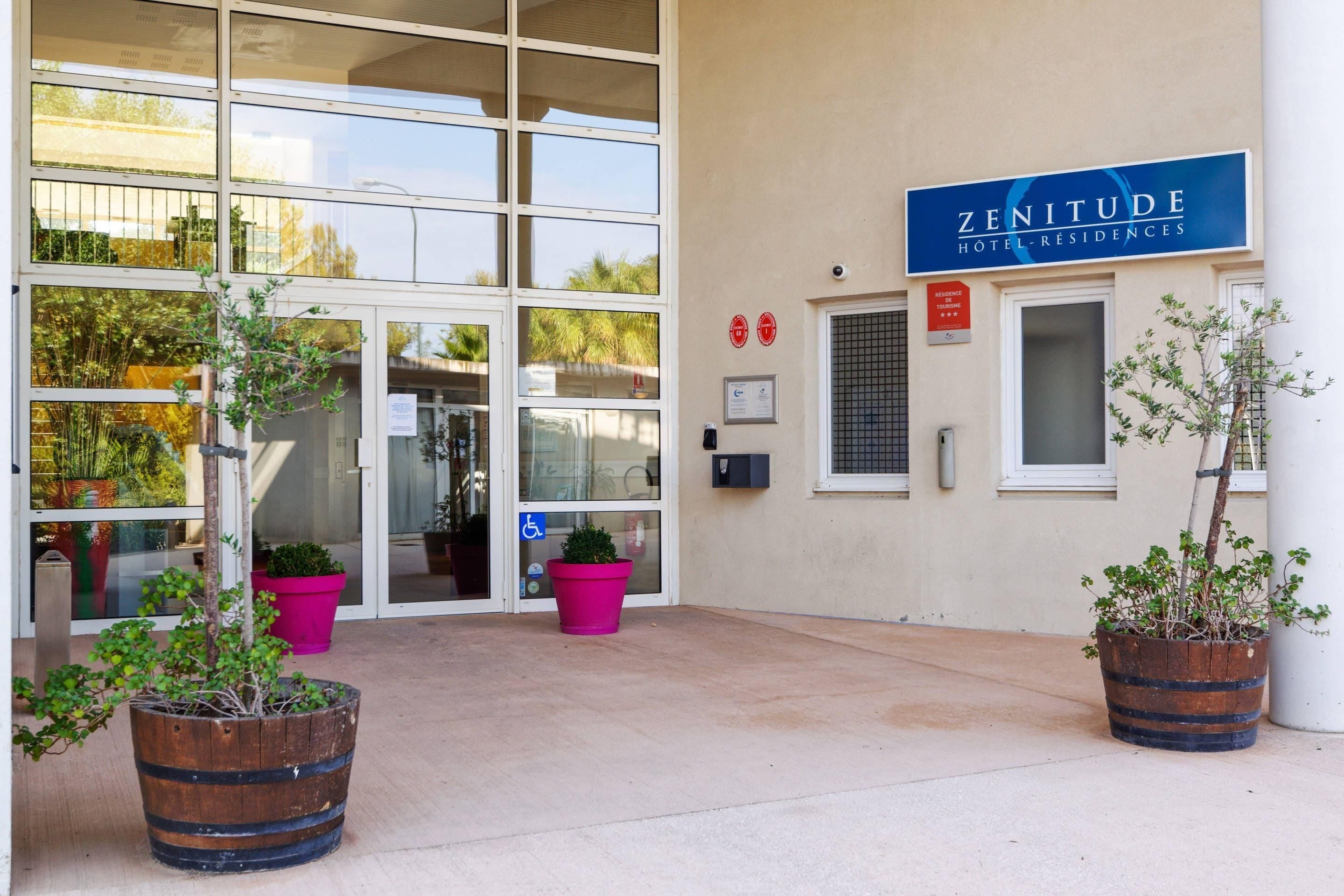 Entrada de hotel com grandes janelas, uma placa com a inscrição 'ZENITUDE HOTEL-RESIDENCES' e plantas em vasos.