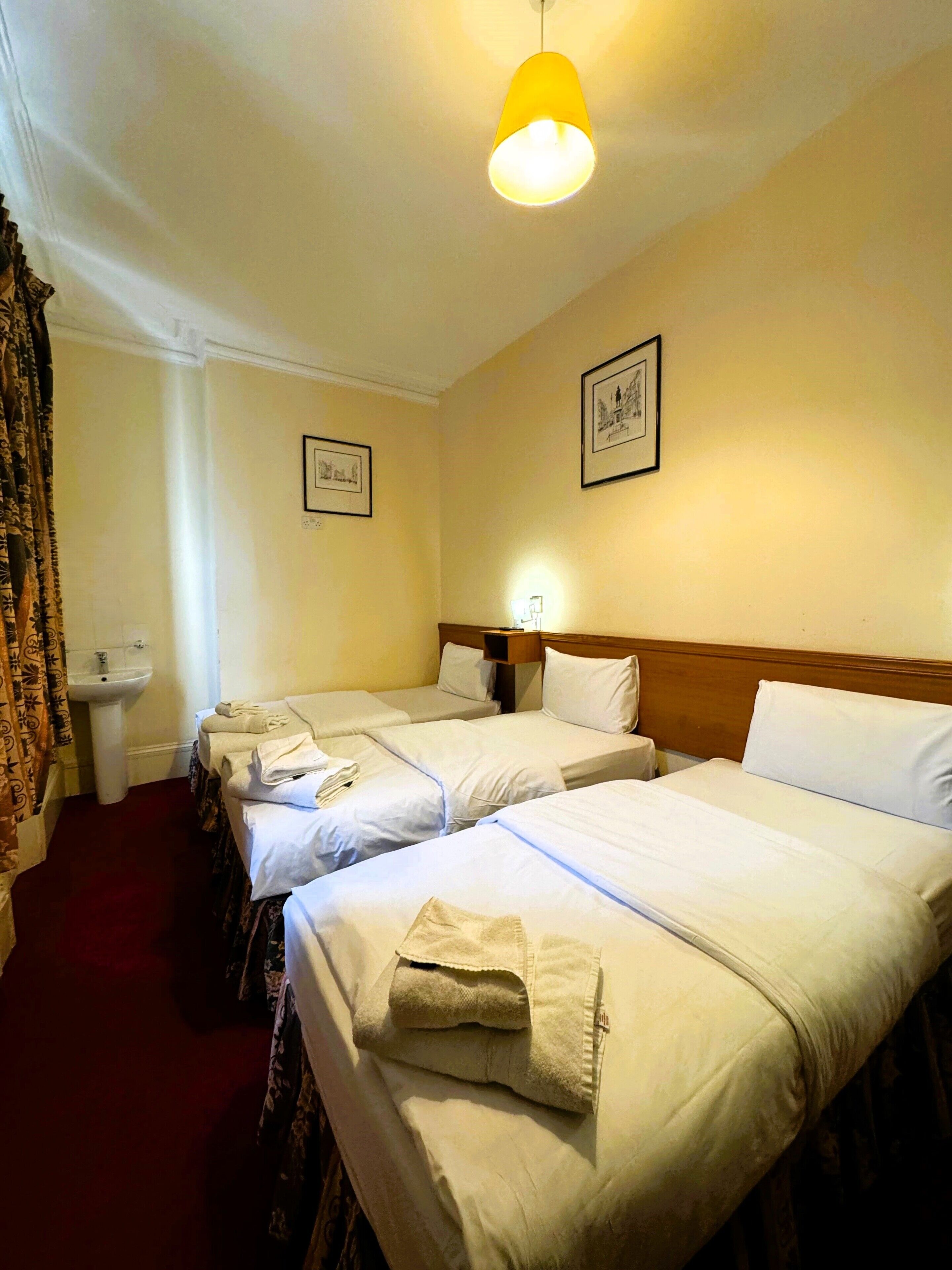 Quarto de hotel com três camas, uma mesinha de cabeceira, uma luminária, uma pia e duas imagens emolduradas na parede.