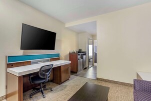 Quarto de hotel com escrivaninha, cadeira, televisão de tela plana, cozinha compacta e vista para o corredor.