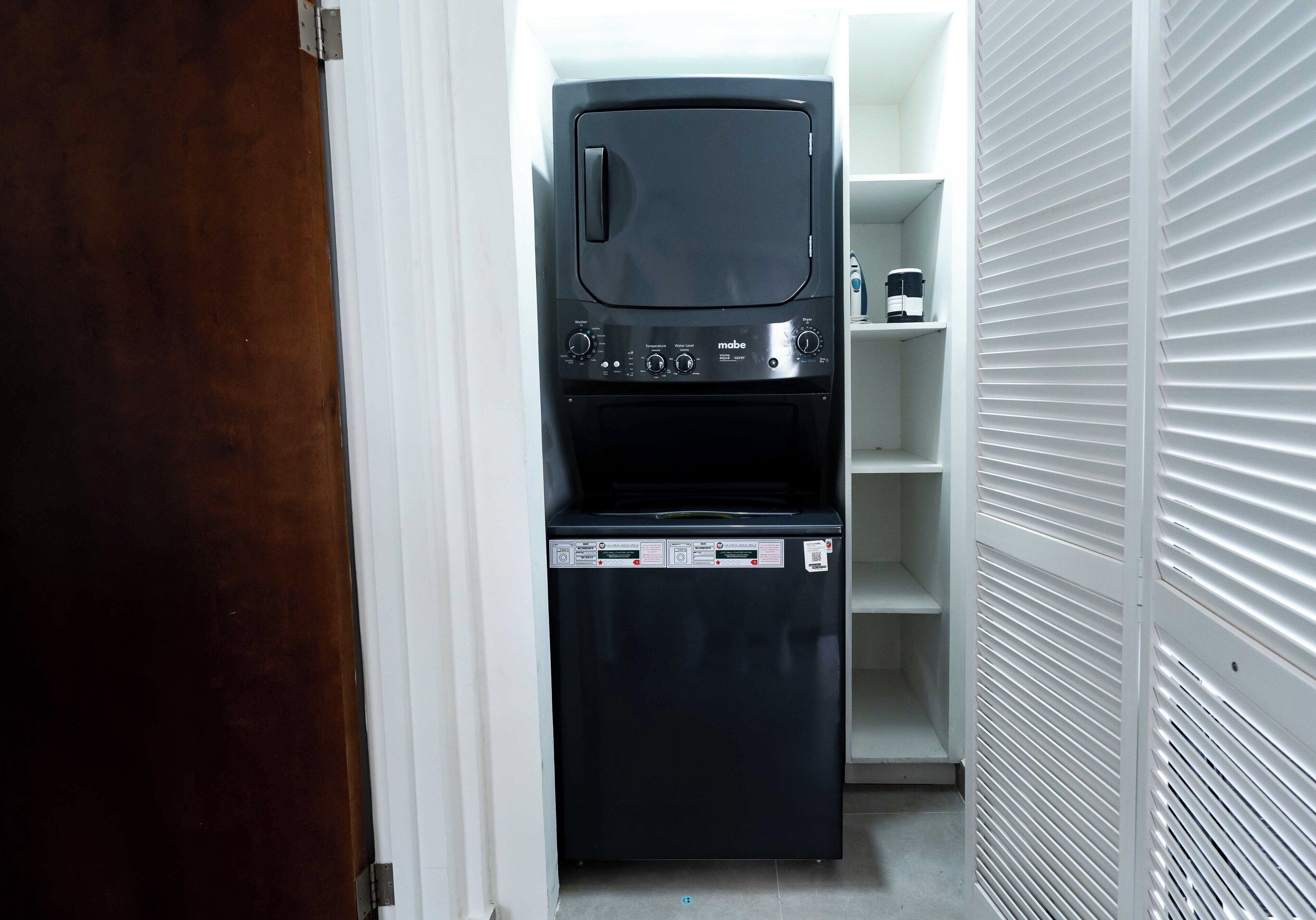 Uma lavanderia com máquina de lavar e secadora, uma prateleira com produtos de higiene pessoal e uma porta branca.
