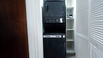 Uma lavanderia com máquina de lavar e secadora, uma prateleira com produtos de higiene pessoal e uma porta branca.