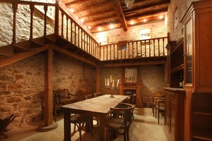 Um interior rústico com uma escada de madeira, paredes de pedra, uma mesa de jantar com cadeiras e uma lareira com um candelabro.