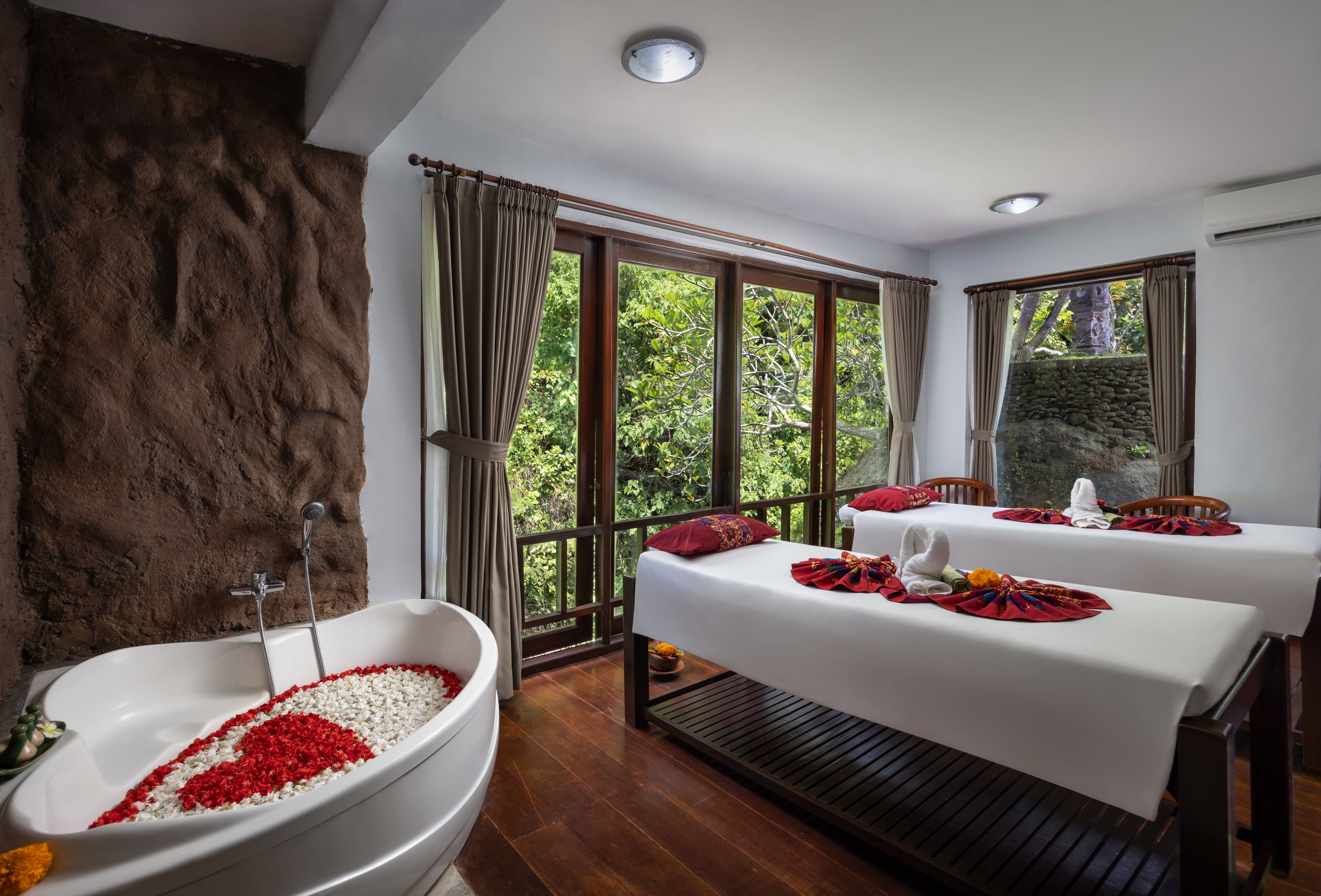 Um espaço de spa com uma banheira cheia de flores vermelhas e brancas, duas mesas de massagem com toalhas e vista para o verde através de grandes janelas.