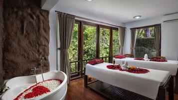 Um espaço de spa com uma banheira cheia de flores vermelhas e brancas, duas mesas de massagem com toalhas e vista para o verde através de grandes janelas.