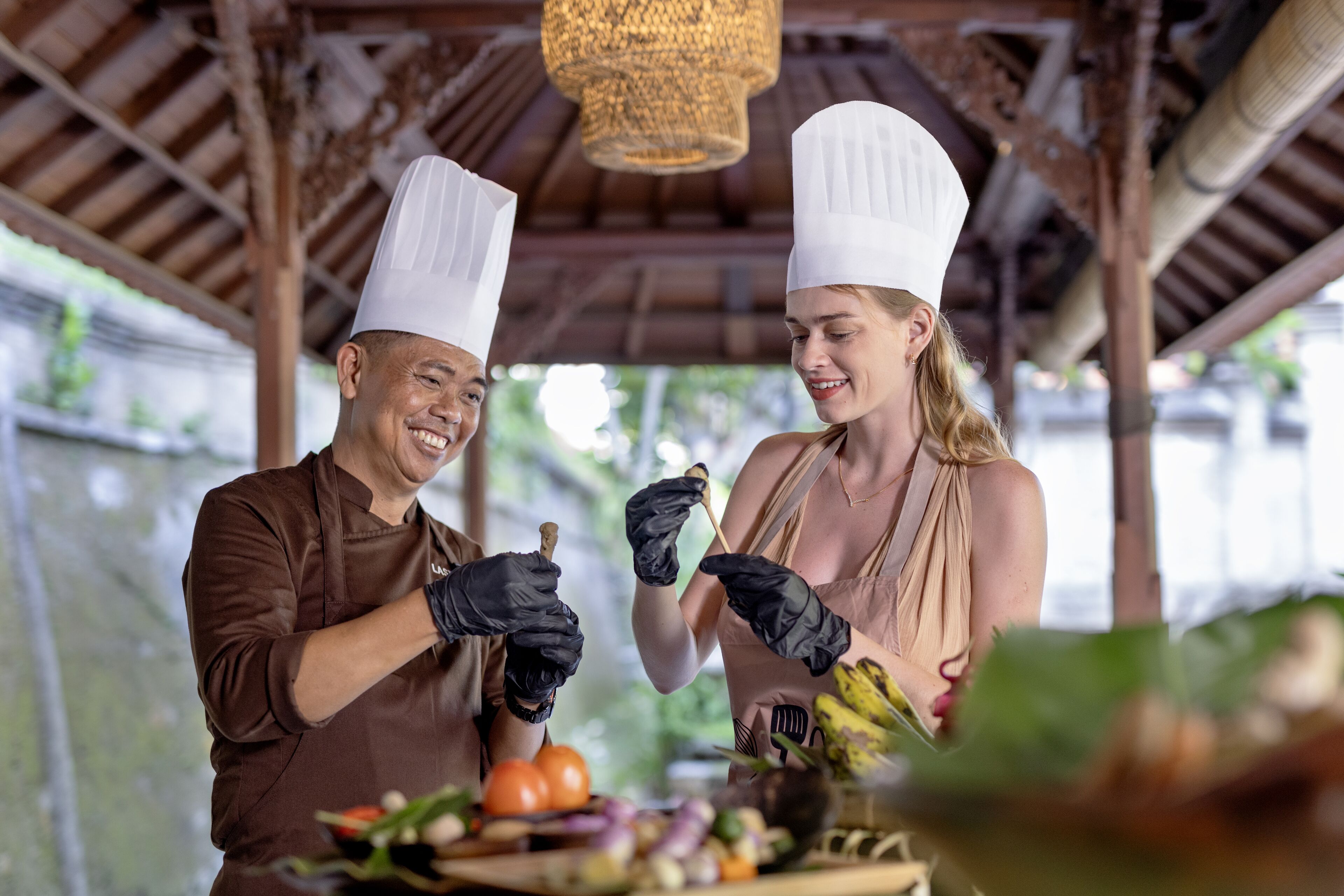 Dois chefs, com chapéus e luvas de cozinha, preparam alimentos em área externa coberta, com estrutura de madeira e luminária pendente.