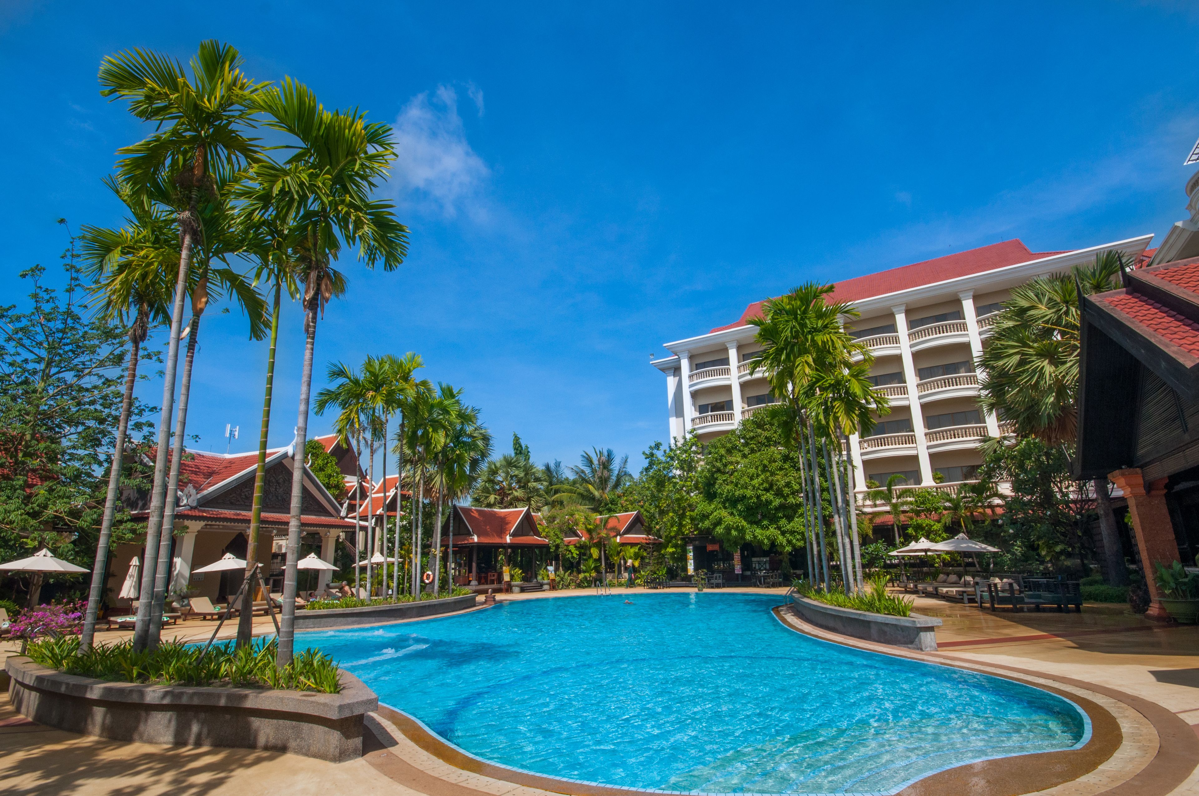 Área de piscina do resort com palmeiras, espreguiçadeiras e um prédio com telhado vermelho.