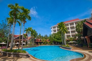 Área de piscina do resort com palmeiras, espreguiçadeiras e um prédio com telhado vermelho.